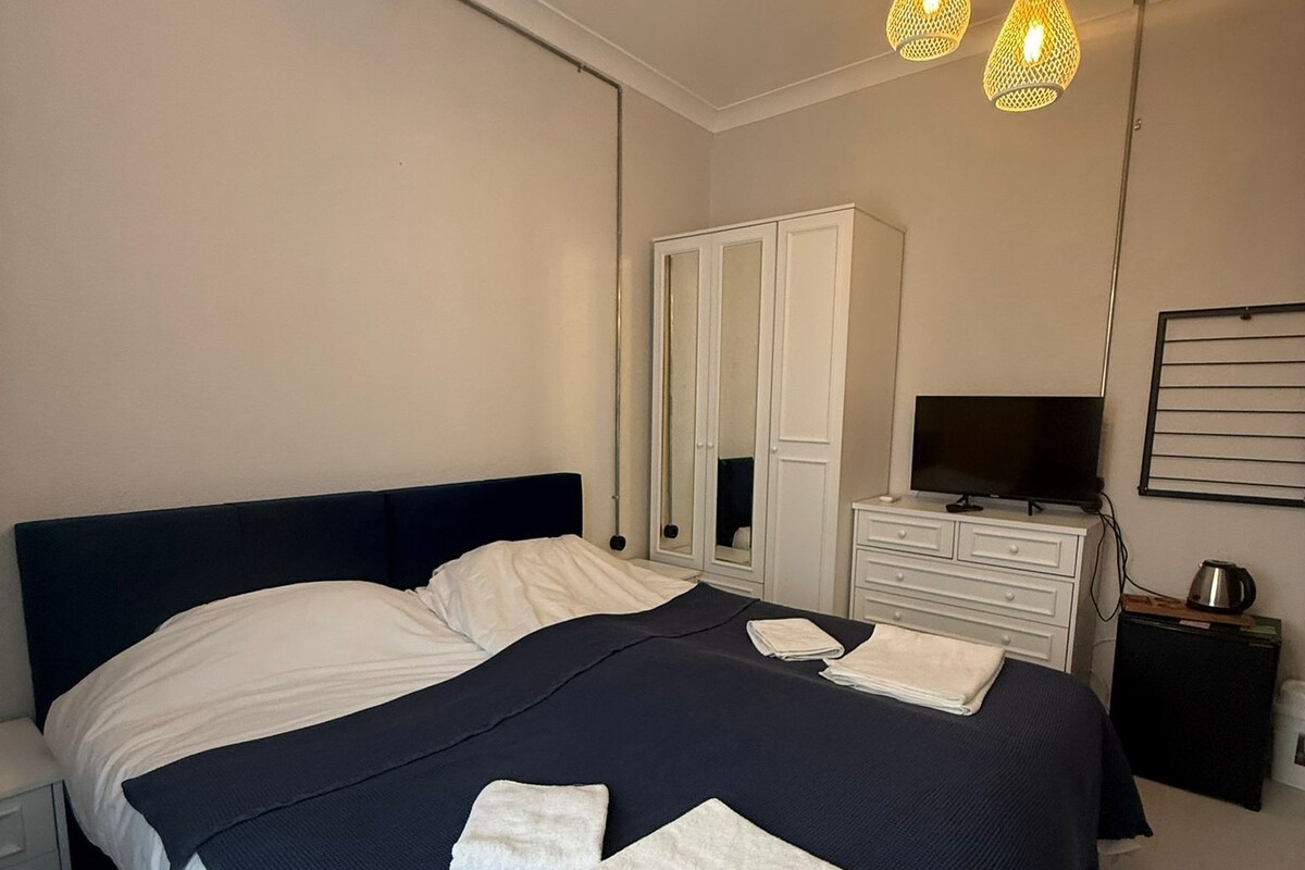 Uma cama bem arrumada com roupa de cama branca e azul marinho, uma pequena mesa de cabeceira com uma chaleira, uma televisão sobre um móvel e um guarda-roupa com espelho.