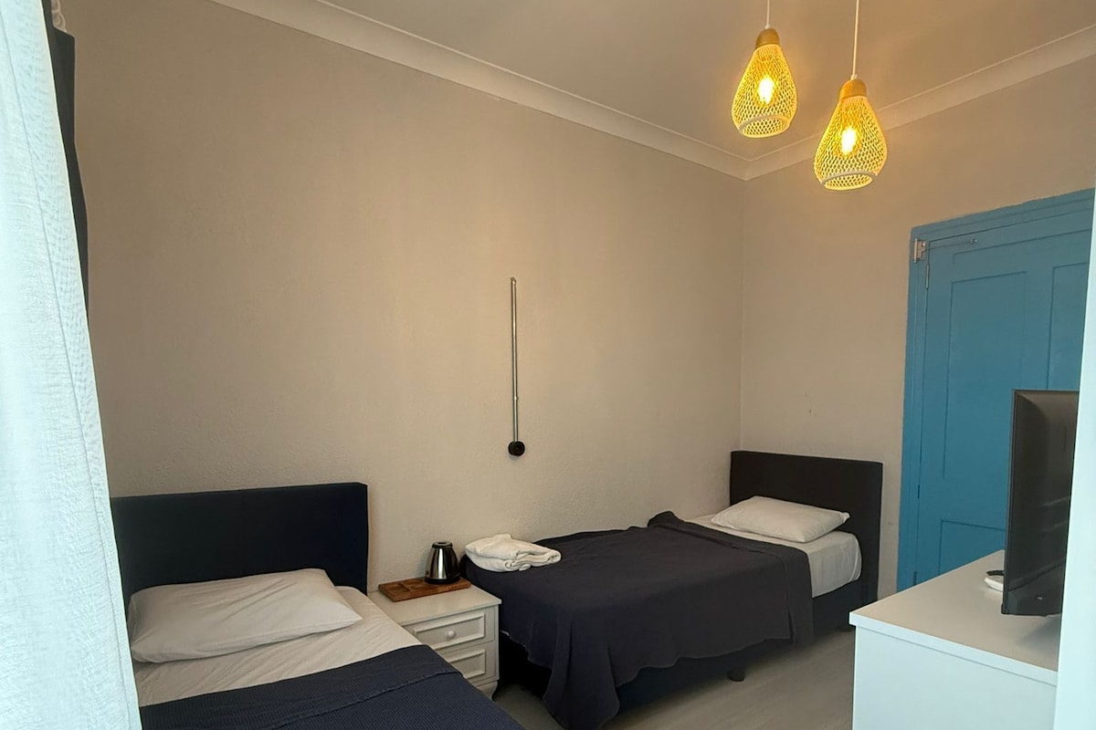 Quarto de hotel com duas camas, uma escrivaninha, televisão e luminárias pendentes.