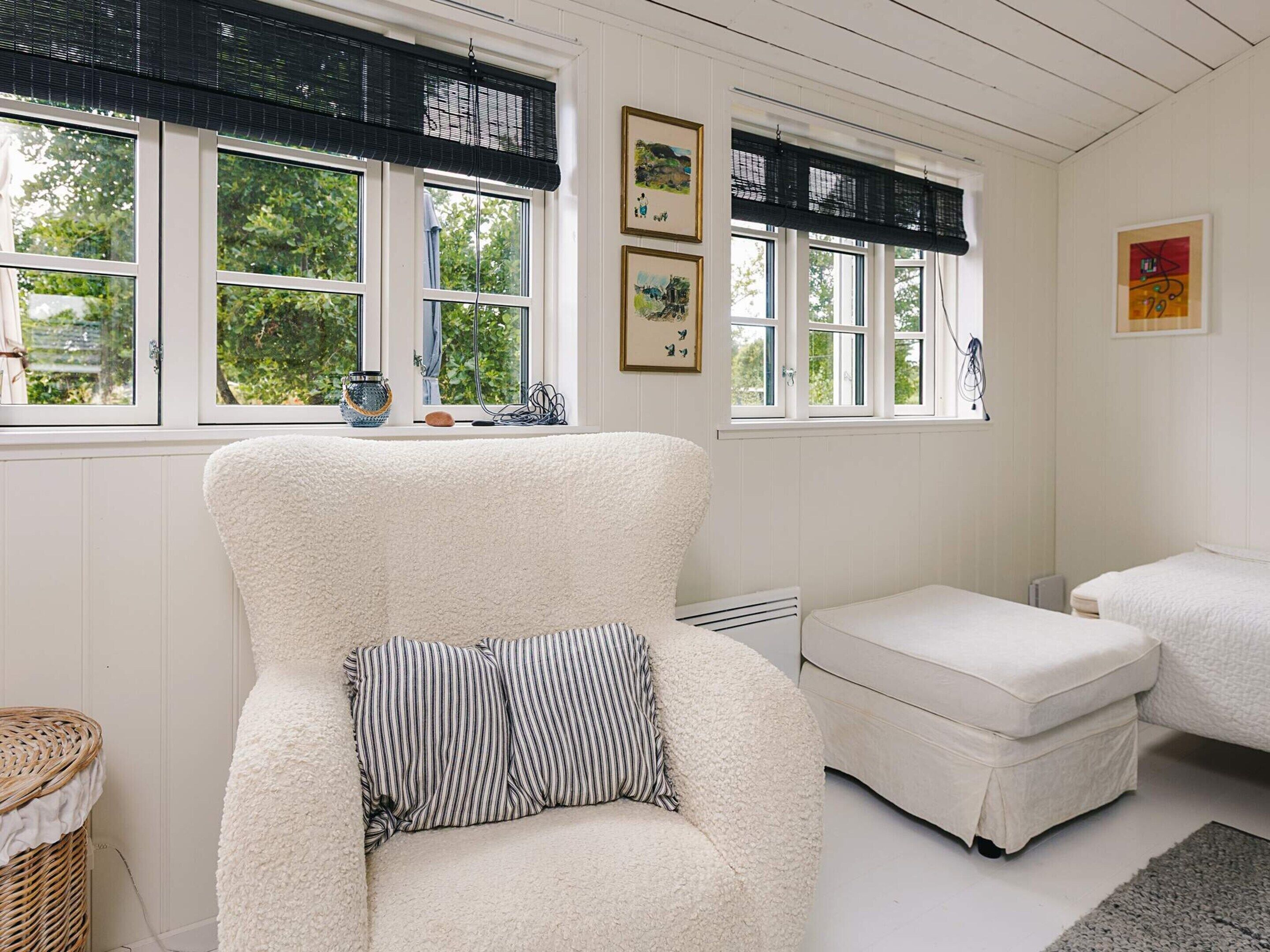 Um quarto com uma poltrona branca, um sofá branco e uma pequena mesa com um vaso e um livro. Há duas janelas com persianas pretas e um quadro na parede.