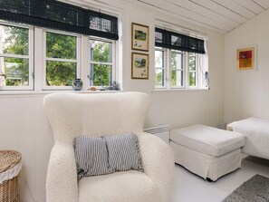Um quarto com uma poltrona branca, um sofá branco e uma pequena mesa com um vaso e um livro. Há duas janelas com persianas pretas e um quadro na parede.