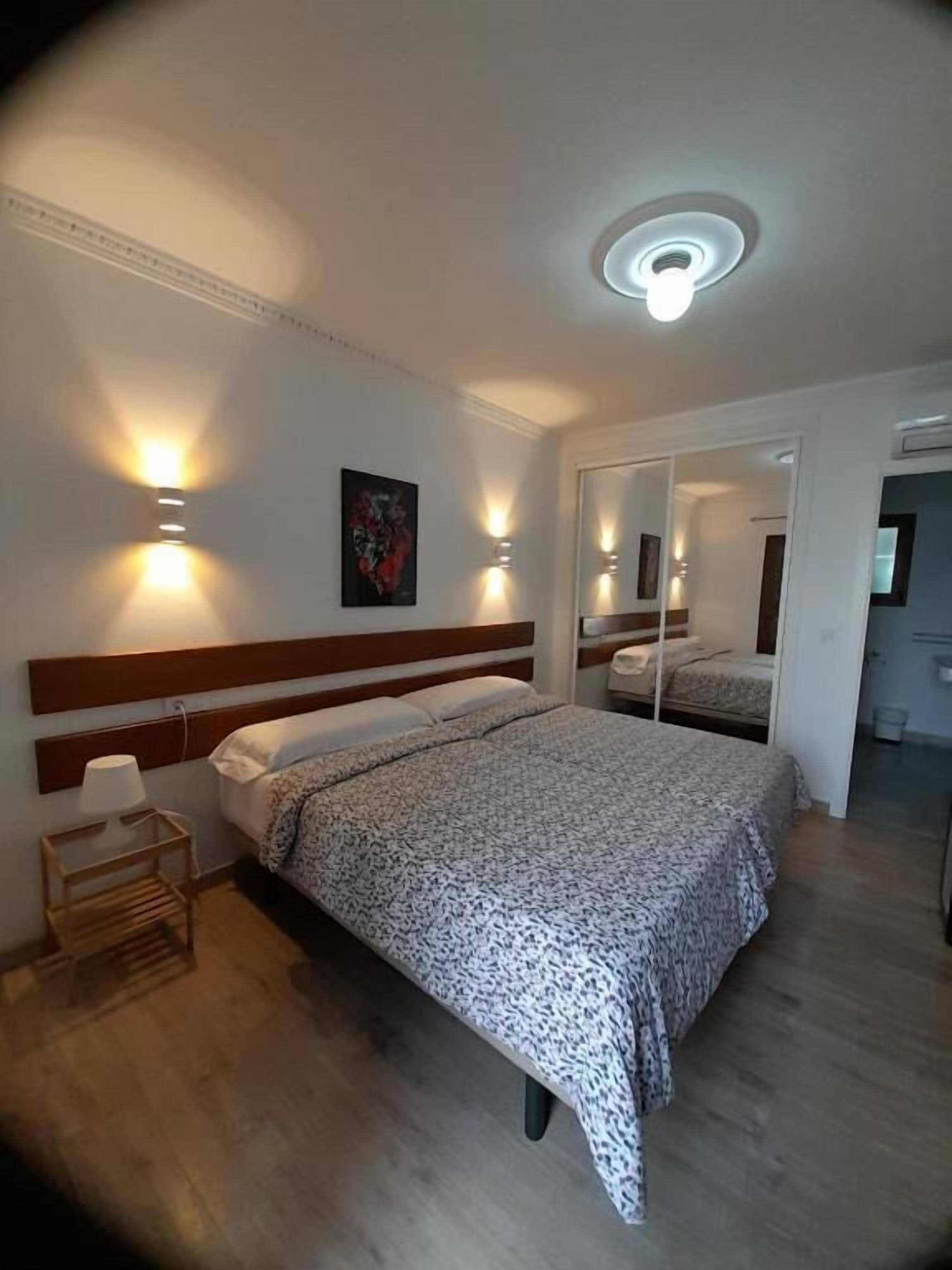 Quarto com cama, luminária de cabeceira, luz na parede e espelho.