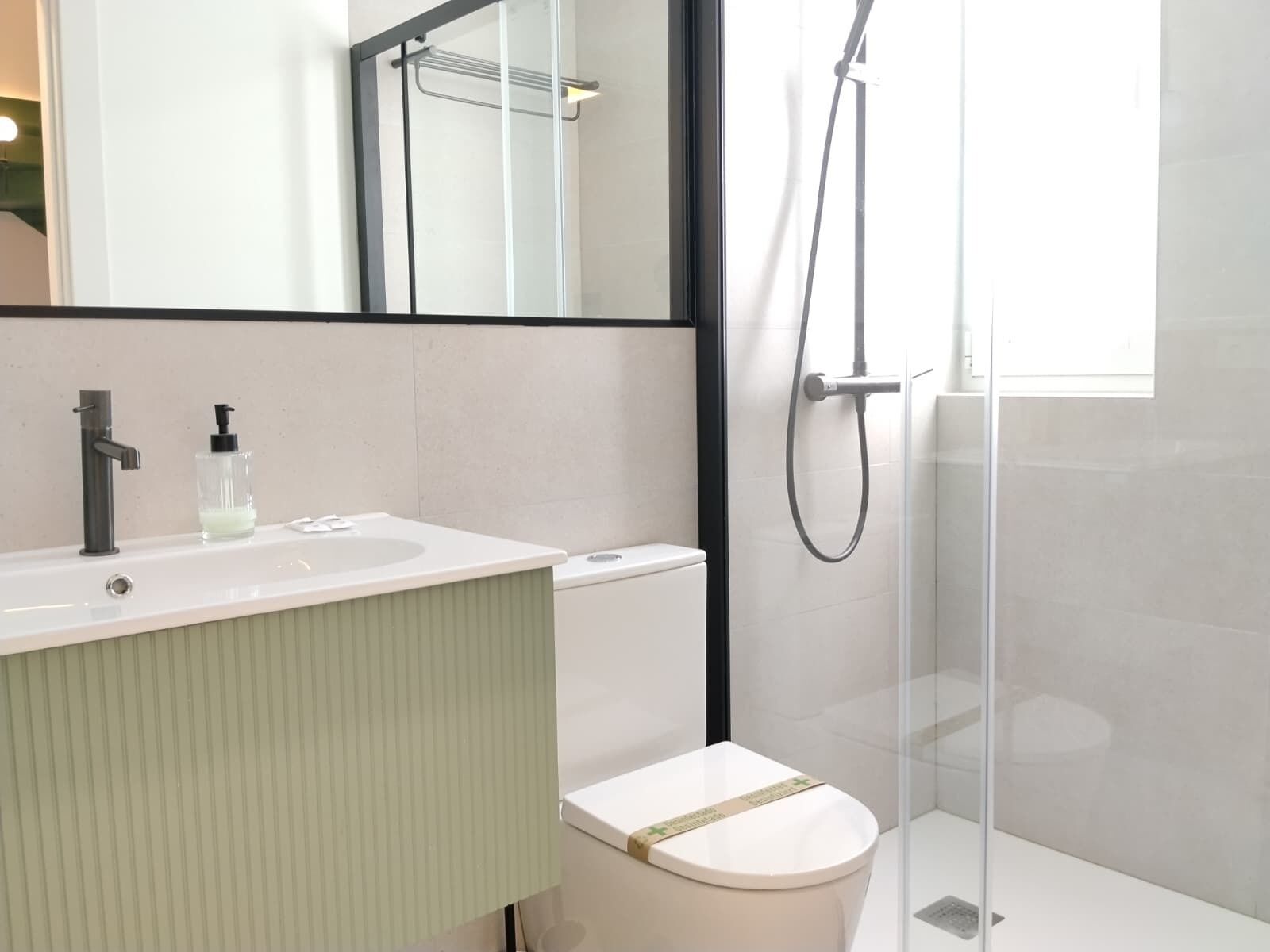 Uma casa de banho moderna com cabine de duche em vidro, lavatório branco com dispensador de sabão e sanita com suporte para toalha verde.