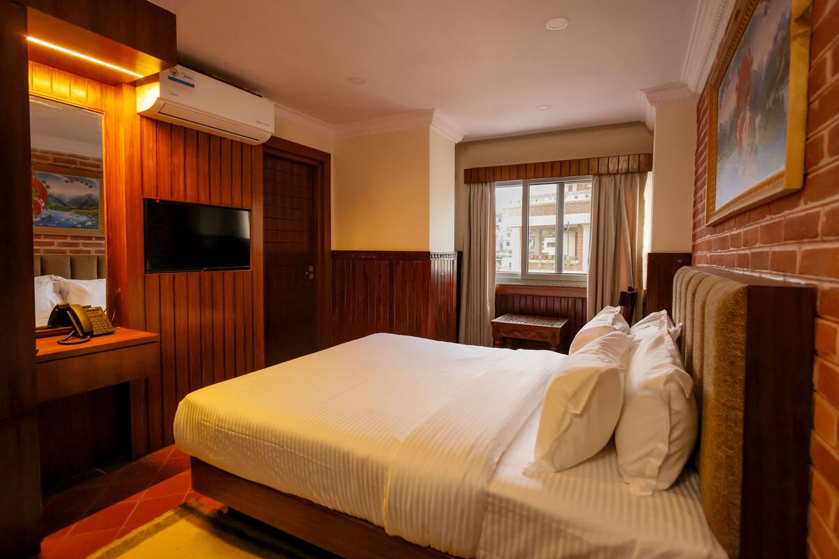 Quarto de hotel com cama, escrivaninha com telefone, televisão e janela com cortinas.