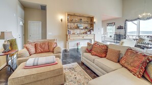 Uma sala de estar ampla com um sofá grande, uma mesa de centro e uma estante de madeira. Uma área de refeições com uma mesa e cadeiras é visível ao fundo.