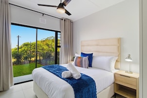 Quarto de hotel com cama, mesa de cabeceira, ventilador de teto e uma janela grande com vista para o verde.