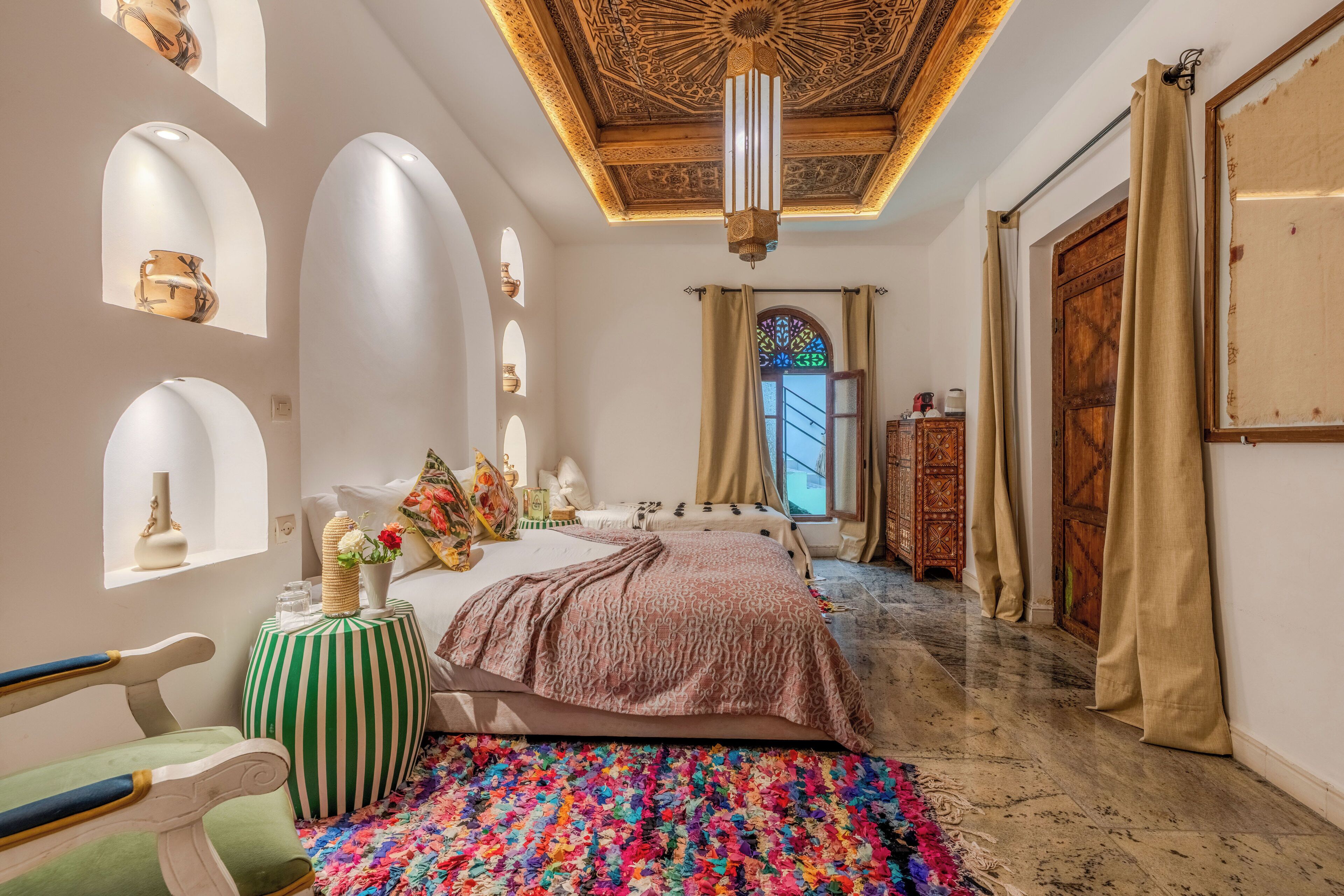 Quarto com cama, um banquinho verde listrado, tapete colorido, armário de madeira e teto decorativo.