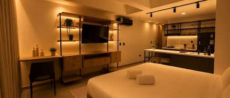 Quarto de hotel moderno com cama, escrivaninha, TV e cozinha compacta.
