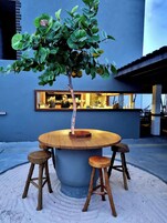 Uma mesa redonda de madeira com bancos, uma árvore e uma parede azul com uma janela.