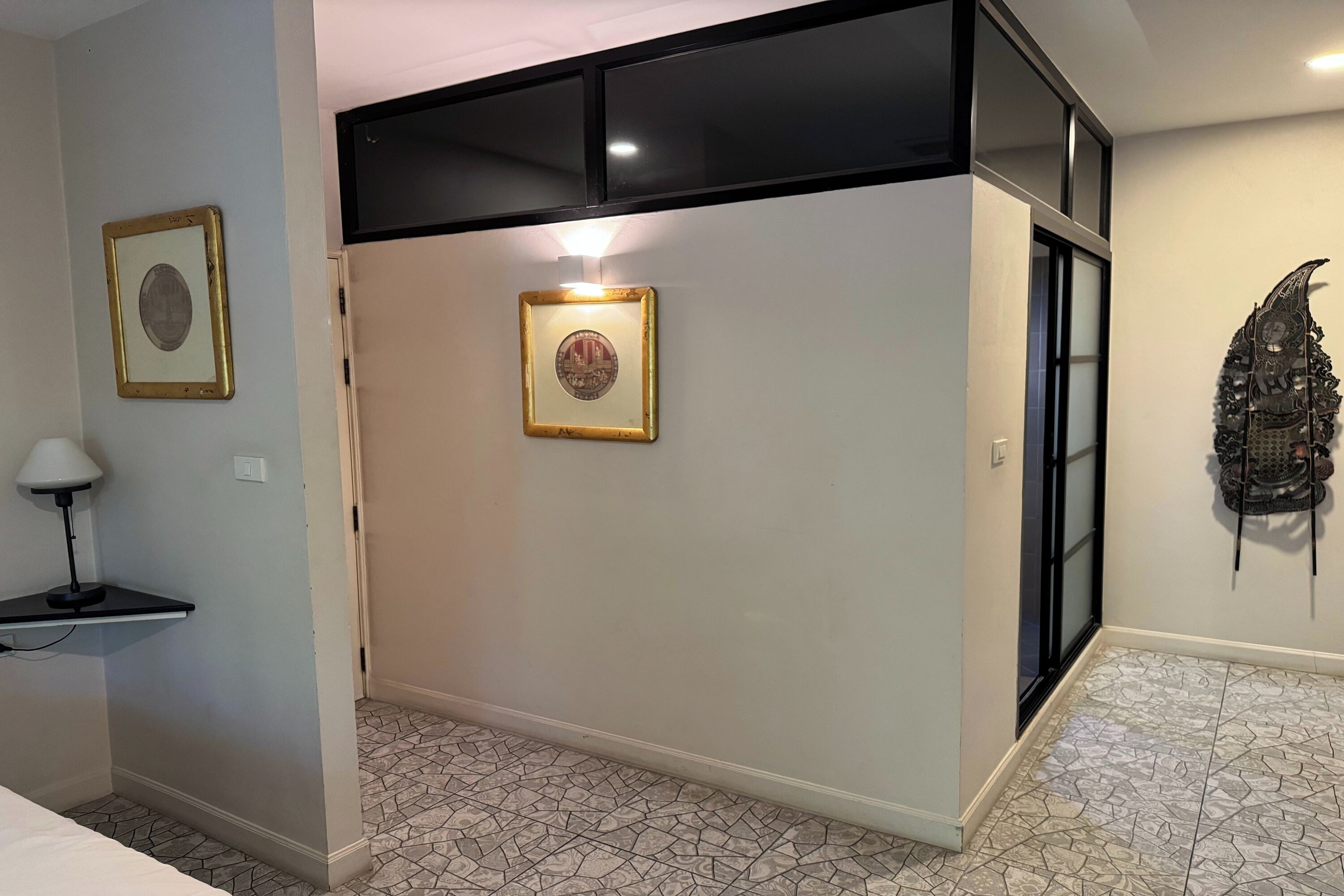 Um corredor com piso estampado, quadros nas paredes, uma escrivaninha com luminária e um objeto decorativo de metal.