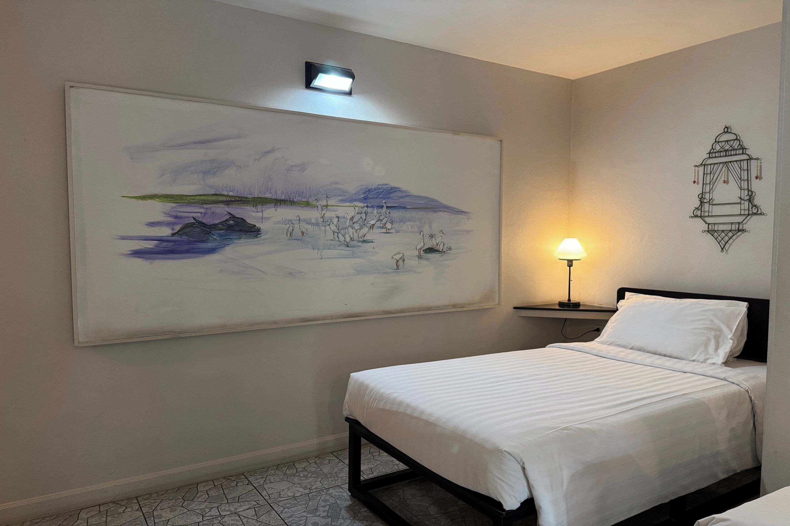 Um quarto com uma cama, uma mesinha de cabeceira, uma luminária na parede e um quadro grande com uma paisagem.