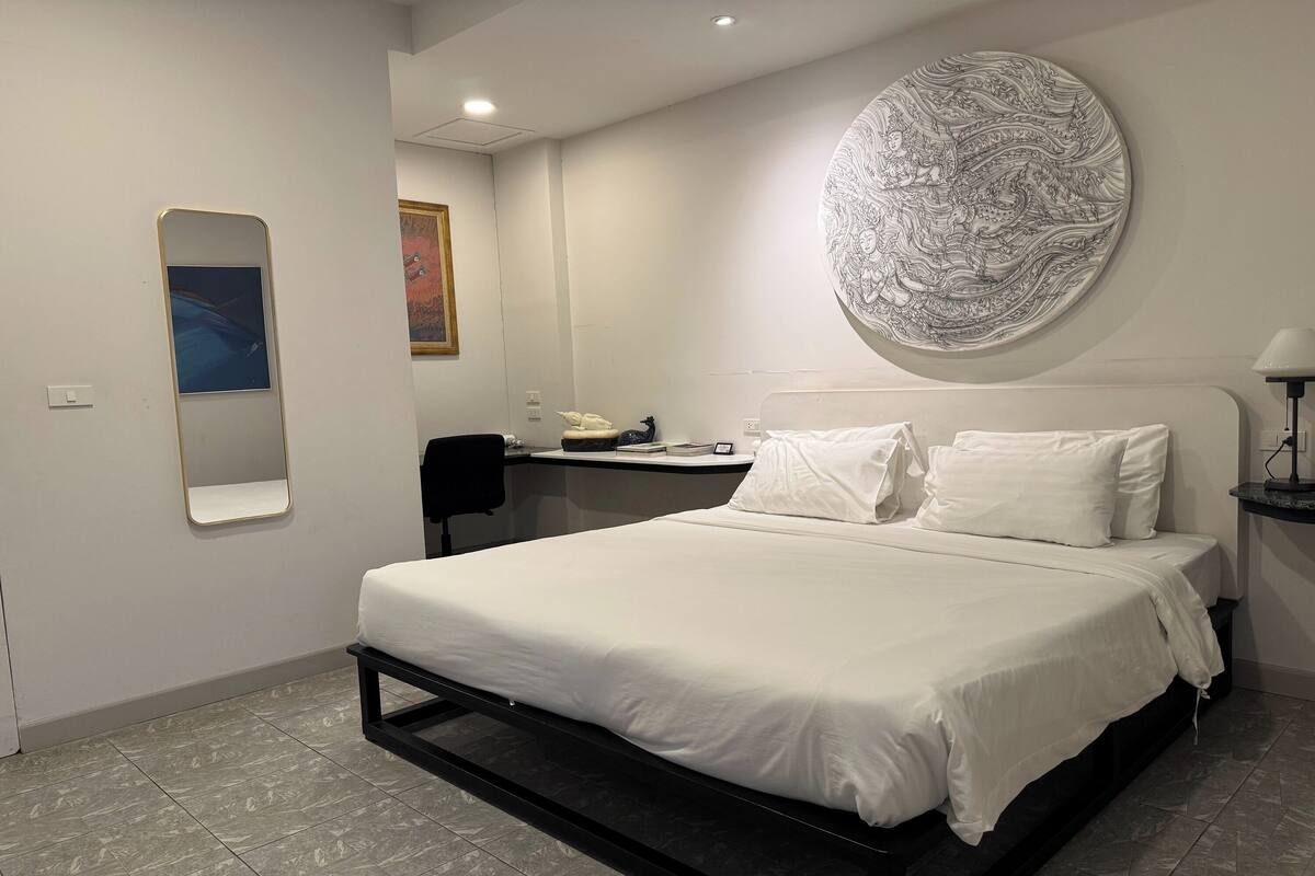 Quarto de hotel moderno com uma cama, uma escrivaninha, uma cadeira, um objeto decorativo redondo na parede e um espelho.