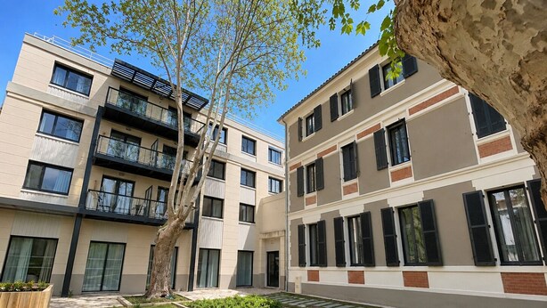 Um edifício residencial com vários andares, varandas, um pátio central com grama e plantas, e uma árvore.