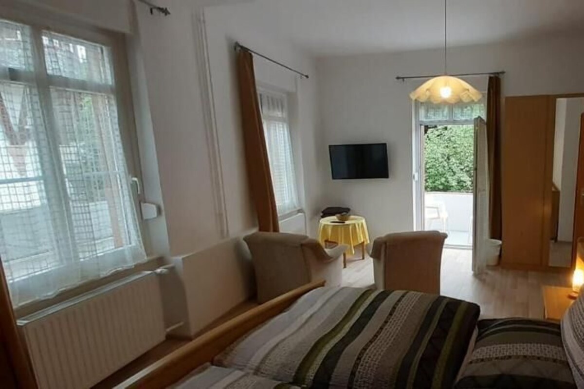 Quarto com cama, cadeira, mesinha, televisão e porta de correr para uma varanda.