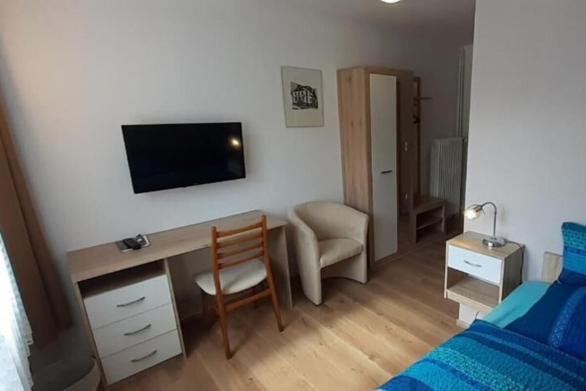 Quarto com cama, escrivaninha, cadeira, televisão, guarda-roupa e mesa de cabeceira.