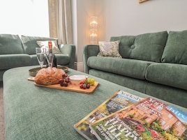 Uma sala de estar com um sofá verde, uma mesa de madeira com pão, uvas e uma garrafa de vinho, e uma revista sobre a mesa.