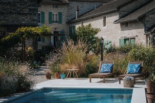 Uma área à beira da piscina, com um edifício de pedra ao fundo, mobiliário de exterior e plantas em vasos.
