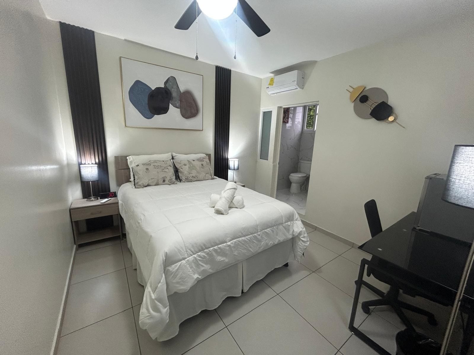 Quarto de hotel com cama, mesa de cabeceira, escrivaninha, cadeira e banheiro visível por uma porta aberta.