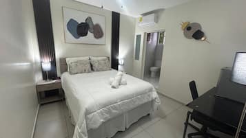 Quarto de hotel com cama, mesa de cabeceira, escrivaninha, cadeira e banheiro visível por uma porta aberta.