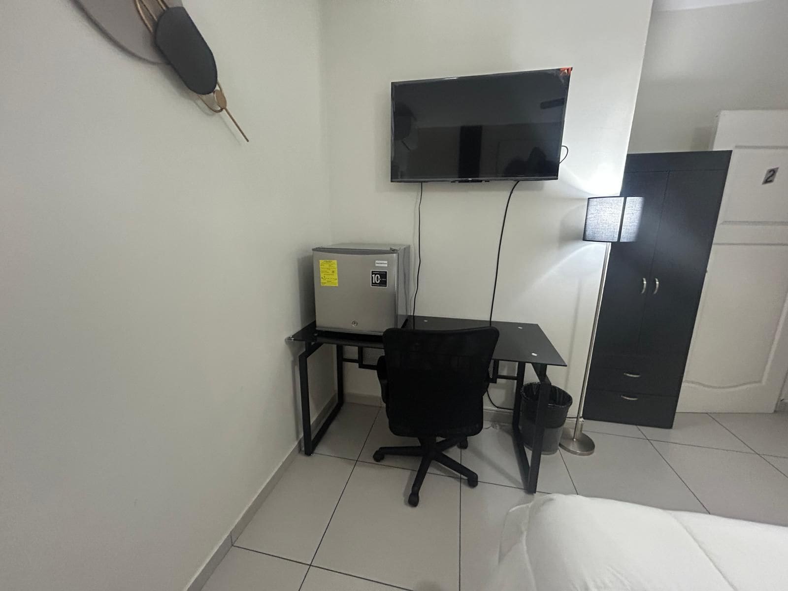 Quarto pequeno com cama, escrivaninha com computador, televisão na parede e guarda-roupa.