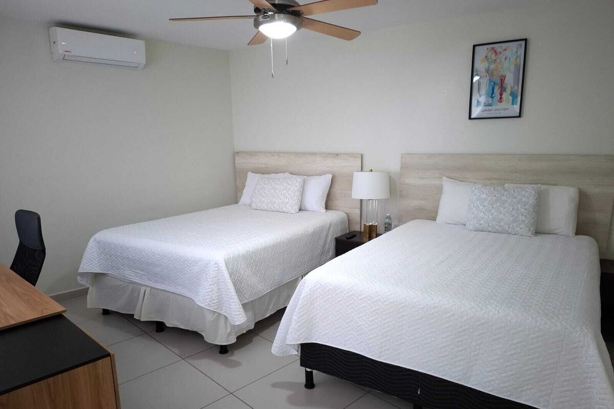 Quarto de hotel com duas camas, um ventilador de teto, ar condicionado, uma escrivaninha e um quadro na parede.