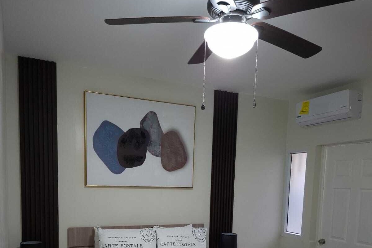 Quarto com cama, ventilador de teto, ar-condicionado fixado na parede, quadro abstrato emoldurado e mesa de cabeceira com abajur.
