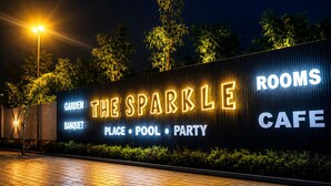 Vista noturna do letreiro 'The Sparkle' com as seções 'Rooms' e 'Cafe', cercado por vegetação e um caminho pavimentado.