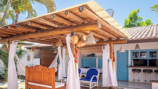 Uma pérgola de madeira com luzes penduradas, uma cadeira azul e um espaço de bar com banquetas.