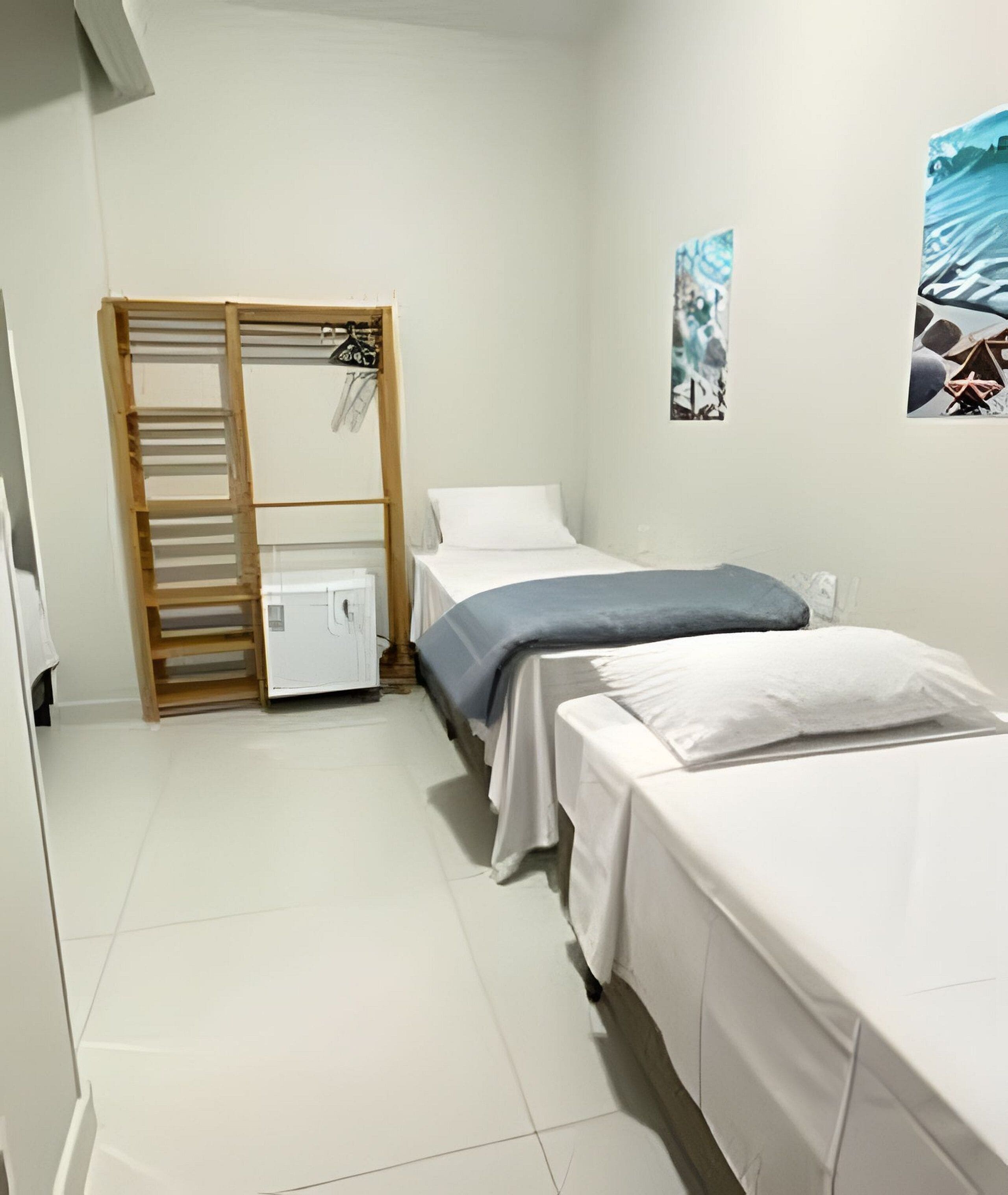 Quarto pequeno e limpo, com duas camas, um guarda-roupa de madeira e uma mesa.