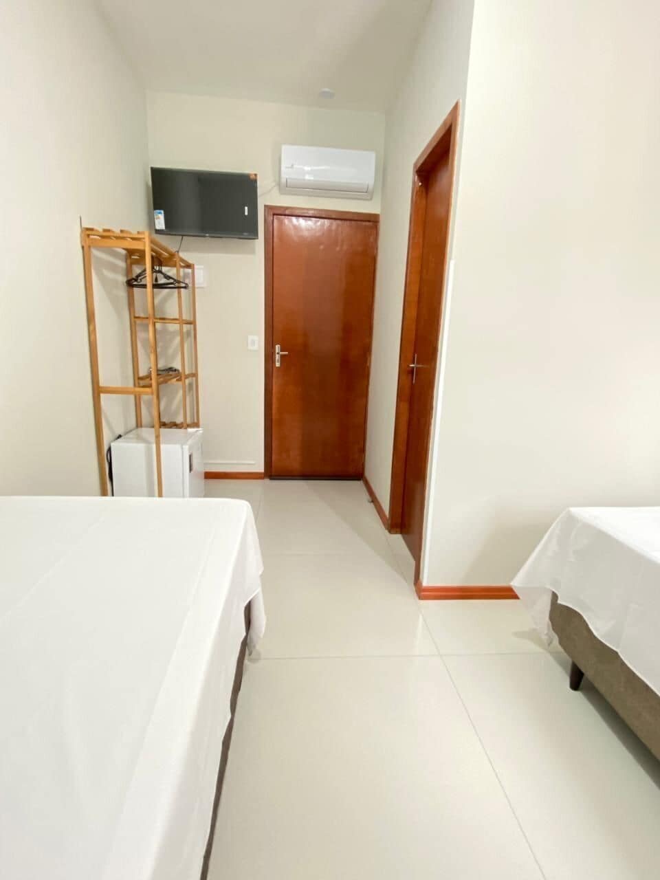 Quarto de hotel estreito com uma cama à esquerda, um armário à direita, uma estante de madeira e uma televisão na parede.