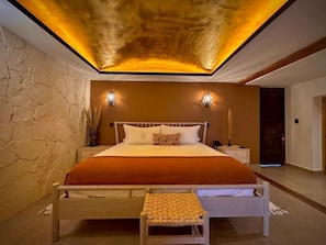 Quarto com cama, mesas de cabeceira, um banquinho de vime e uma parede de pedra.