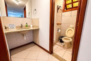 Casa de banho com lavatório, sanita e chuveiro. Há um sinal no lavatório e um rodapé de porta de madeira.