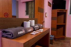 Quarto de hotel com cabeceira de madeira, um banco com toalhas, um cesto de lixo e um guarda-roupa.
