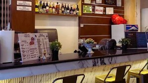 Uma área de bar com balcão, banquetas, um quadro de cardápio e uma estante com diversas garrafas.