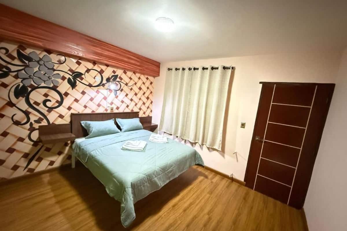 Um quarto com uma cama, uma mesa de cabeceira, parede com padrão geométrico, janela com cortinas e uma porta.