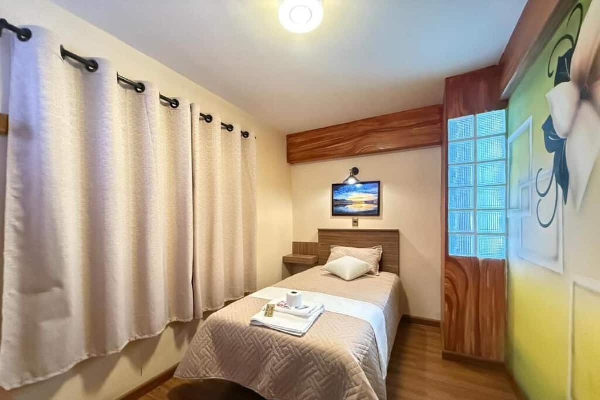 Cama bem arrumada com colcha, uma pequena mesa de cabeceira com abajur, uma janela com persianas e um quadro fixado na parede.