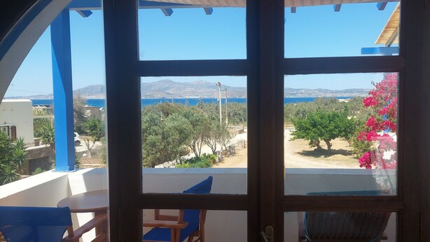 Quarto com uma janela grande que oferece vista para o mar, árvores e montanhas ao longe.