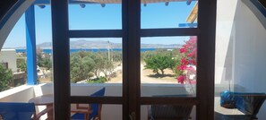 Quarto com uma janela grande que oferece vista para o mar, árvores e montanhas ao longe.