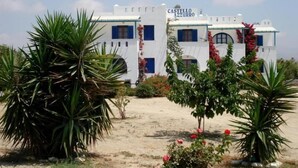 Um edifício branco com a sinalização 'CASTELLO AZZURRO', rodeado por vegetação e um chão de areia.