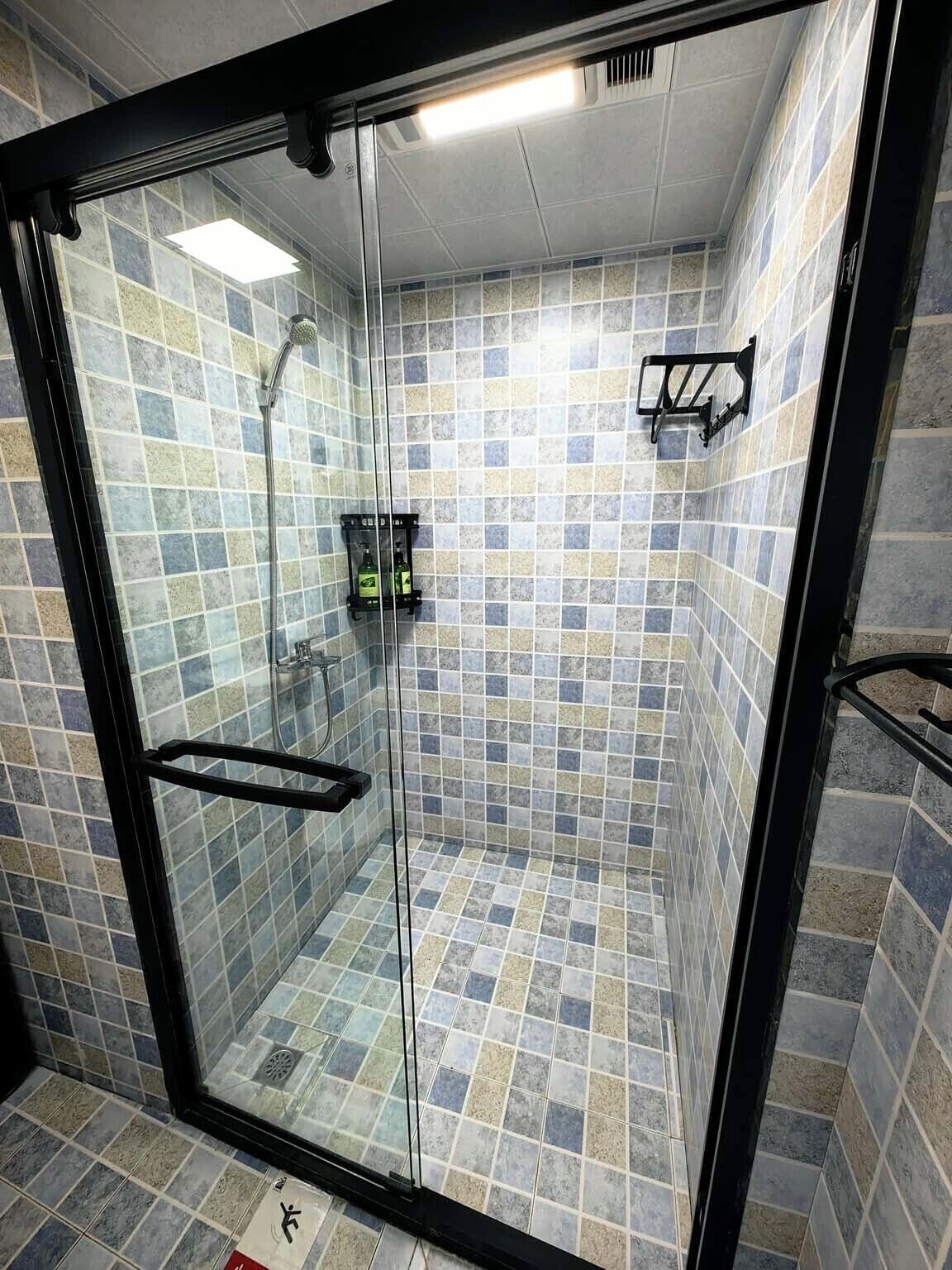 Área de chuveiro com paredes revestidas de azulejos e moldura preta, protegida por vidro.