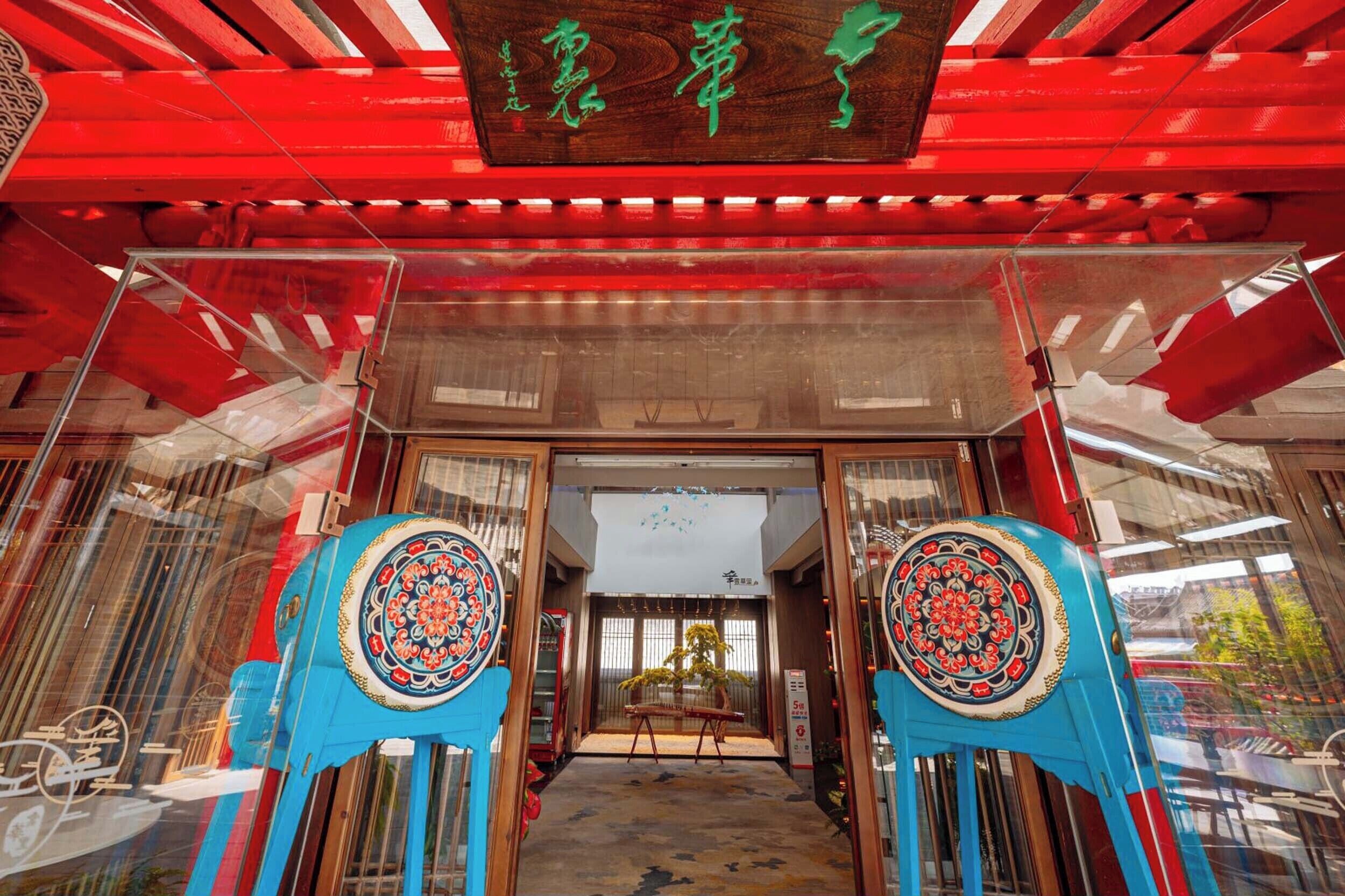 Uma entrada tradicional japonesa com uma placa de madeira, dois pilares decorativos azul e vermelho, e um caminho central que leva a um aposento.