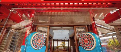 Uma entrada tradicional japonesa com uma placa de madeira, dois pilares decorativos azul e vermelho, e um caminho central que leva a um aposento.
