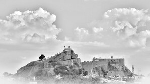 Uma fortaleza histórica com uma formação rochosa imponente, uma torre de vigia e uma muralha fortificada.