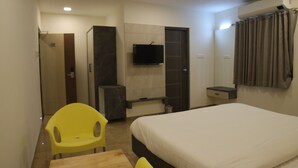 Quarto de hotel com cama, televisão fixada na parede, mesinha e cadeira amarela.