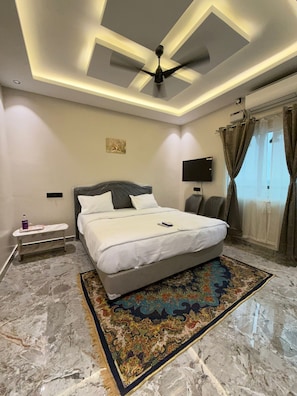 Quarto com cama, ventoinha no teto, televisão, mesa de cabeceira, janela com cortinas e tapete estampado.