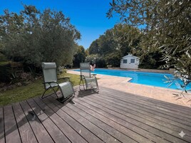 Um deck de madeira com duas espreguiçadeiras voltadas para uma piscina, cercado por árvores e um pequeno galpão.