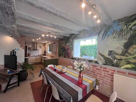 Uma sala de jantar com uma mesa comprida, cadeiras, uma televisão e uma cozinha ao fundo.