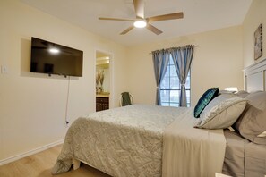 Quarto com cama, ventilador de teto, TV fixada na parede, janela com cortinas e mesa de cabeceira com abajur.