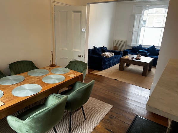 Uma sala de jantar com mesa de madeira e cadeiras verdes, uma sala de estar com sofá azul e uma mesa de centro, e uma porta branca.