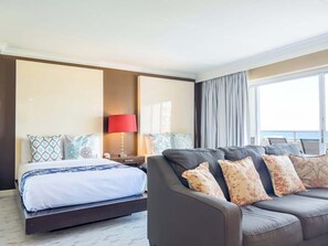 Quarto de hotel com cama, sofá, mesa de cabeceira com abajur e uma janela grande com cortinas.