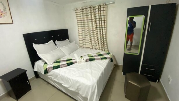 Um quarto com uma cama, mesa de cabeceira, guarda-roupa, espelho e cortina.
